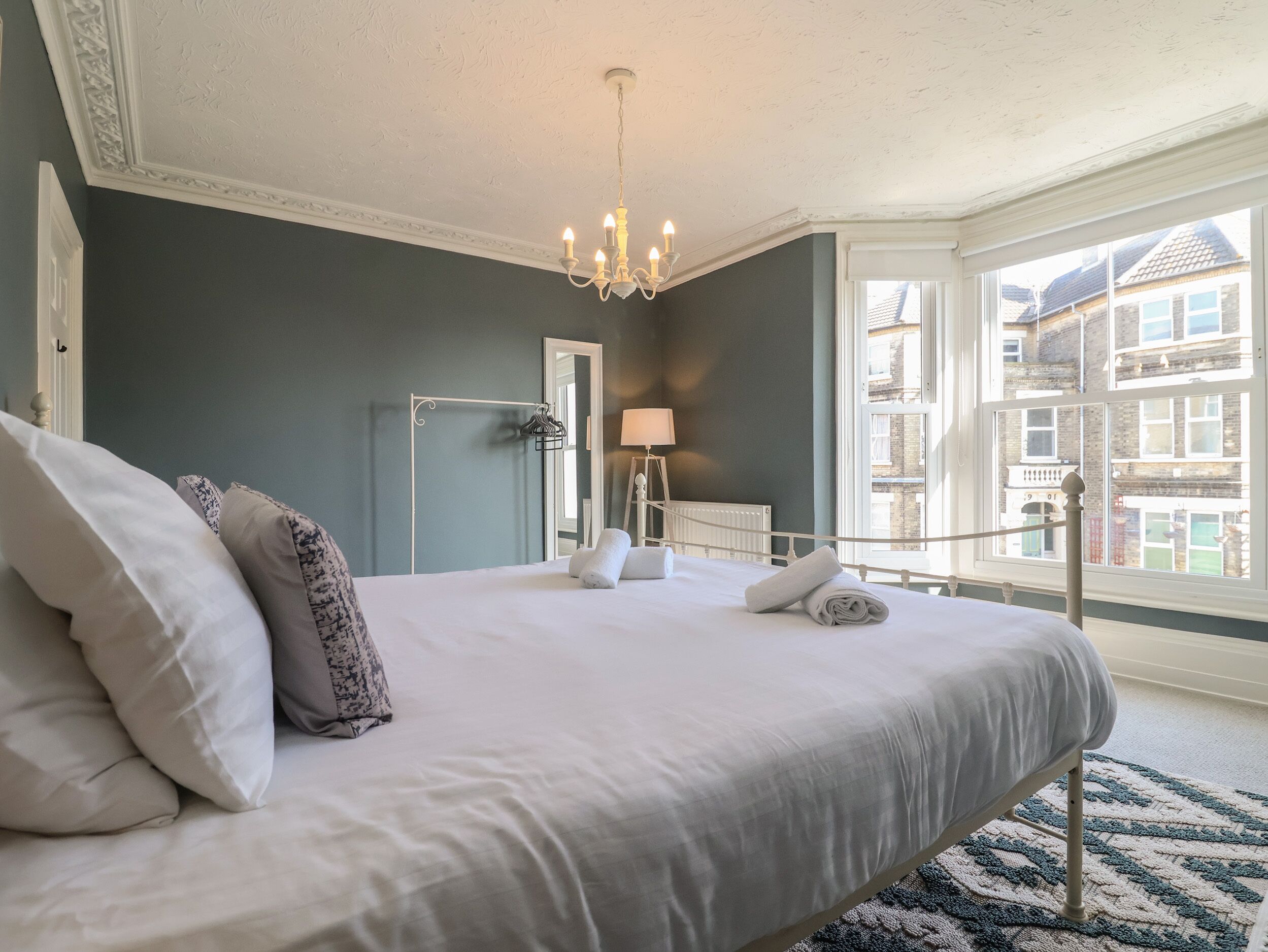 Uma cama bem arrumada com lençóis brancos e almofadas decorativas, um candelabro, um espelho e vista para edifícios através de uma grande janela.