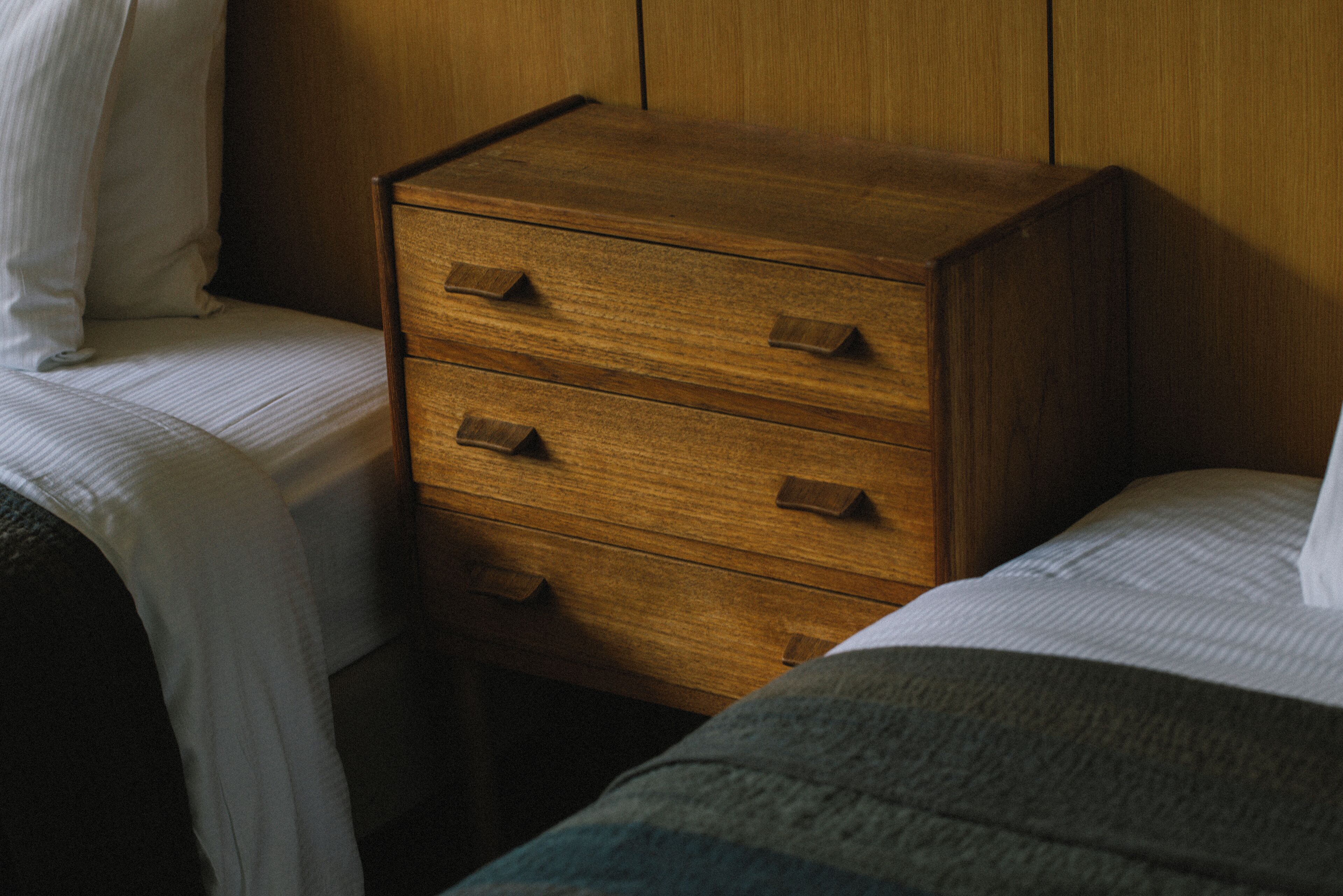 Uma cômoda de madeira com três gavetas, uma cama com lençol branco e uma cabeceira de madeira.