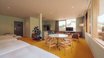 Quarto de hotel com duas camas, área de refeições com mesa e cadeiras, televisão e janela com vista para o exterior.