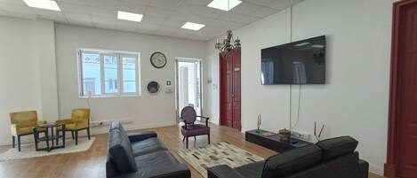 Uma sala de estar ampla com um sofá modular preto grande, uma poltrona amarela e uma poltrona roxa. Há uma TV de tela plana na parede, um lustre e um relógio.