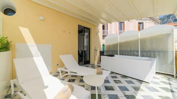 Uma área de estar ao ar livre iluminada pelo sol, com poltronas brancas, uma mesa redonda pequena e um sofá branco.