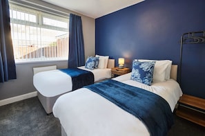 Quarto de hotel com duas camas, uma janela com cortinas, um radiador e uma mesa de cabeceira com abajur.