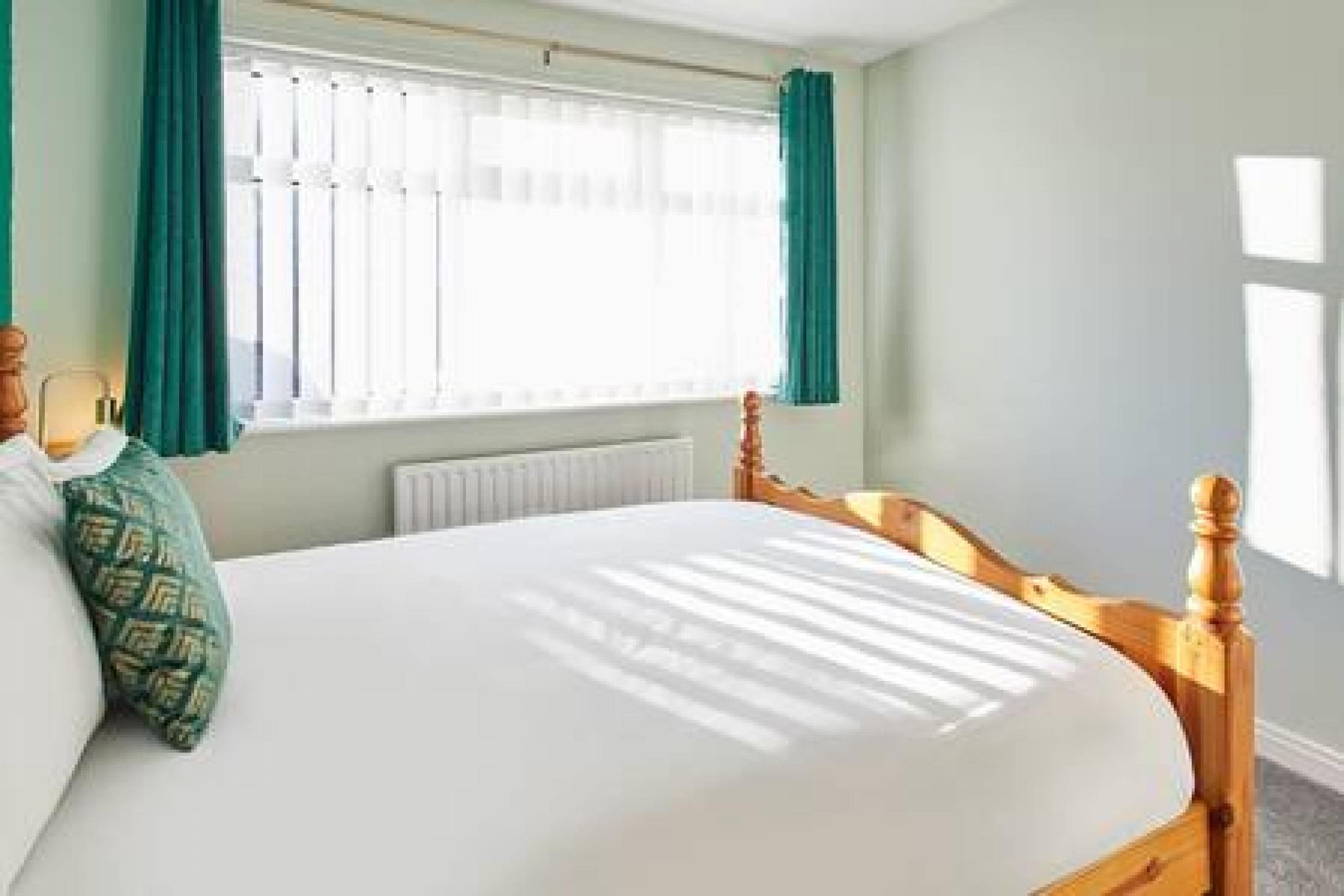 Uma cama bem arrumada com cabeceira de madeira, janela com persianas brancas e cortinas verdes.