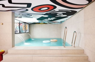 Uma piscina interior moderna com água cristalina, duas barras de apoio de metal e um mural colorido no teto.