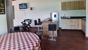 Quarto com cama estampada em xadrez, pequena escrivaninha com cadeiras, televisão, geladeira e cozinha com armários de madeira.