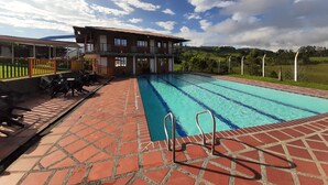 Uma piscina com deck ladrilhado, um edifício com várias janelas e varandas, e vista para colinas verdes sob um céu parcialmente nublado.
