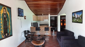 Uma sala de estar ampla com teto de madeira, piso ladrilhado, sofá preto, mesa de centro de madeira e cozinha ao fundo.