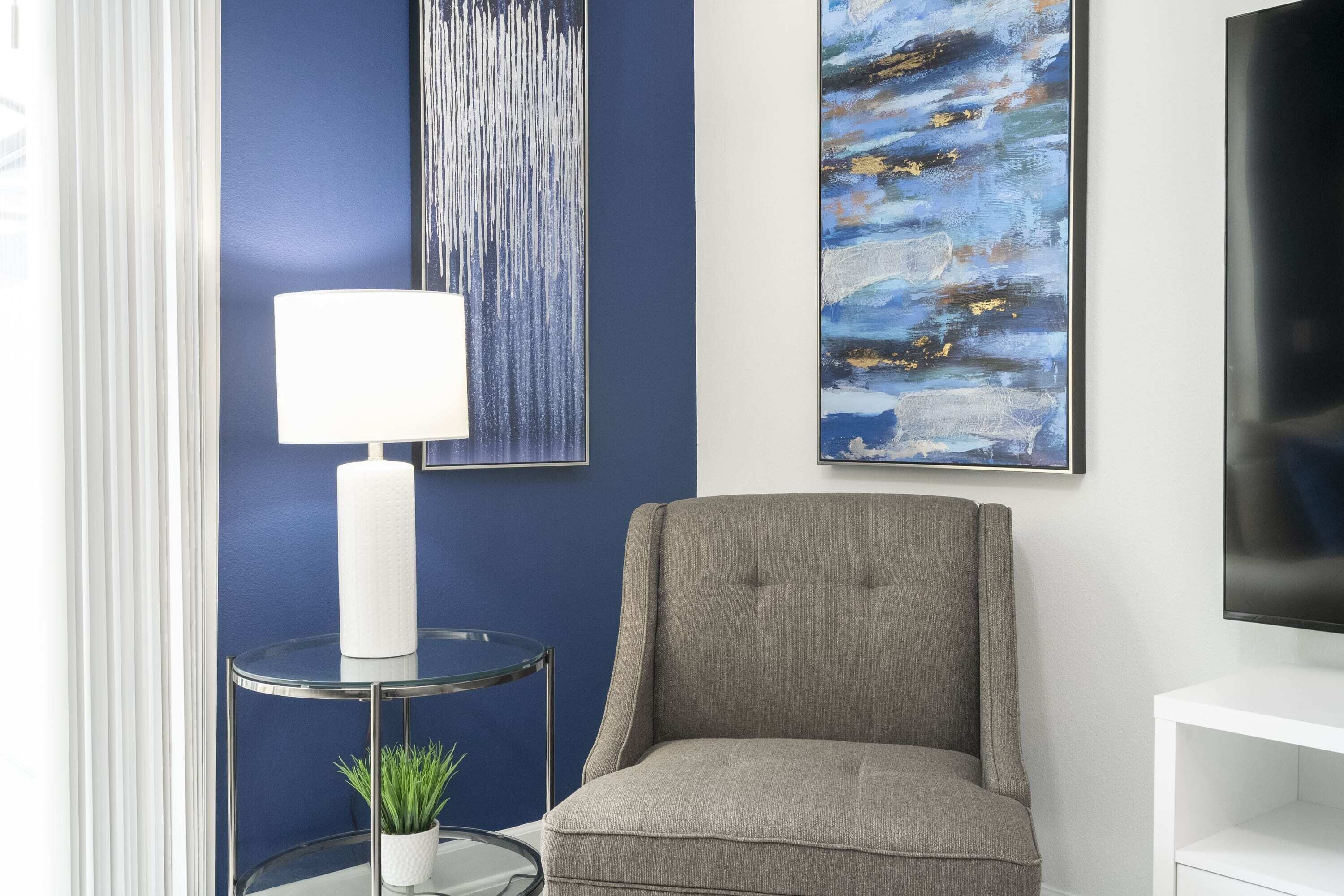Uma sala de estar moderna com uma poltrona cinza, uma mesa lateral de vidro com abajur e um quadro na parede.