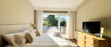 Quarto de hotel com uma cama grande, televisão, cómoda de madeira e varanda com vista para a cidade.