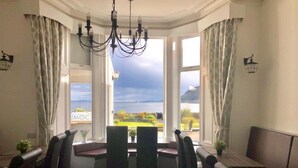 Uma sala de jantar com vista para o mar, um candelabro e uma mesa posta para seis pessoas.