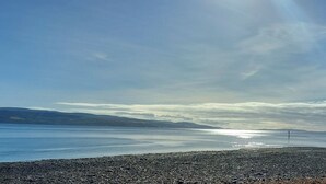 Paisagem costeira com praia de seixos, águas calmas e montanhas ao longe.