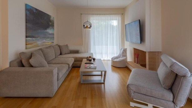 Uma sala de estar com sofá, poltrona e mesa de centro. Há um quadro na parede, uma televisão instalada acima da lareira e uma janela com cortinas leves.