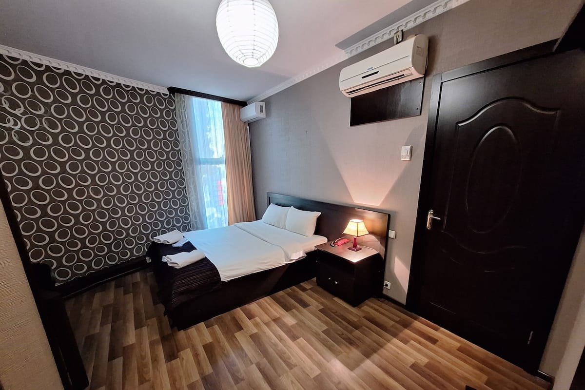 Quarto de hotel com cama, mesa de cabeceira, luminária, aparelho de ar condicionado e uma porta.