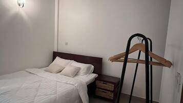 Uma cama bem arrumada com lençóis brancos, uma mesinha de cabeceira de madeira com duas gavetas, uma luminária fixada na parede e um cabide de madeira sobre um suporte.