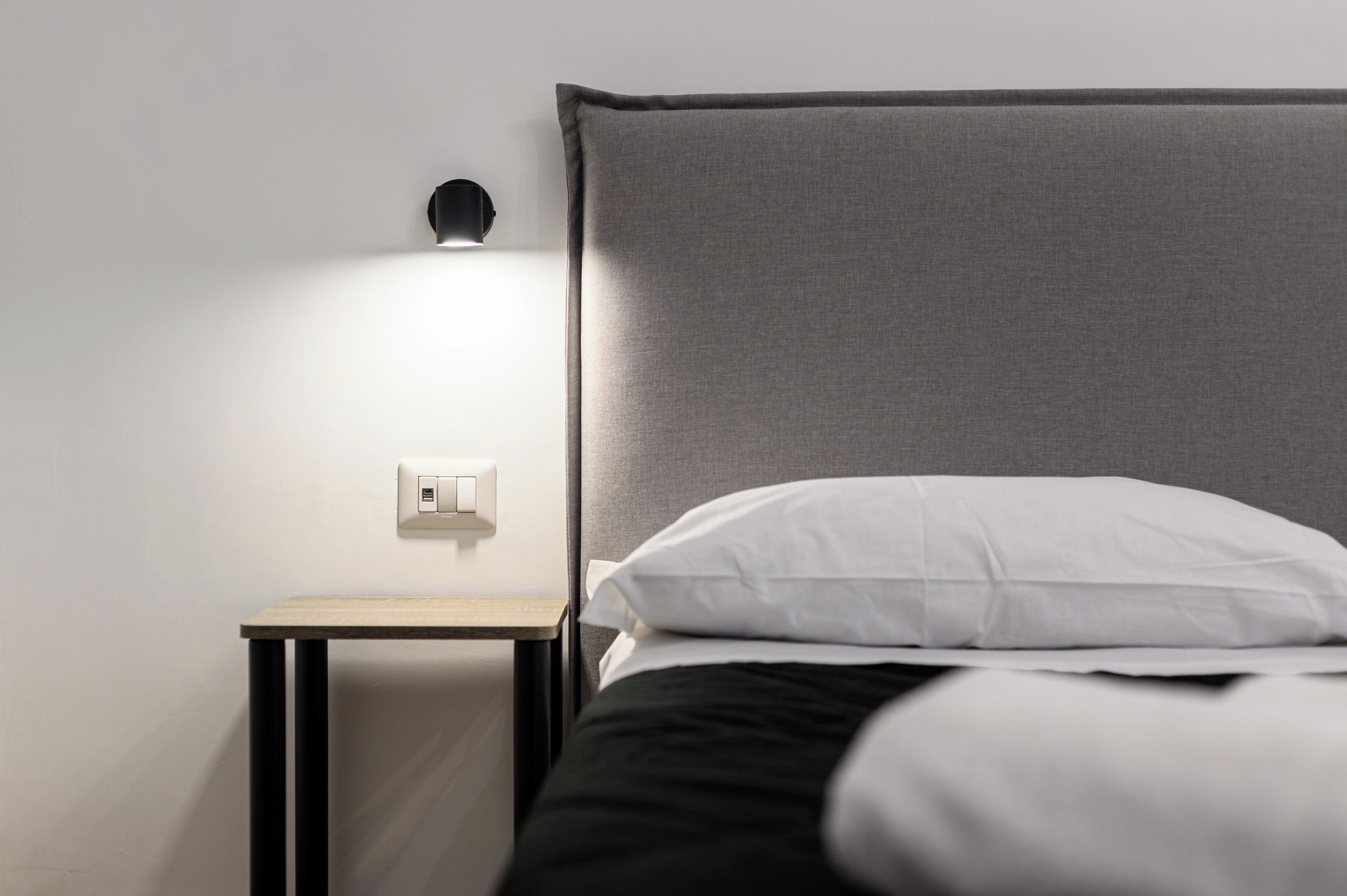 Cama bem arrumada com roupa de cama branca e cabeceira cinza, uma pequena mesa de cabeceira com abajur e uma tomada na parede.