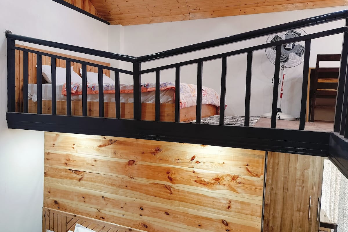 Uma escada de madeira com corrimão preto leva a um quarto com cama e ventilador.