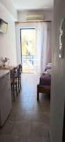 Quarto de hotel compacto com cozinha compacta, uma cama com colcha roxa, uma mesa pequena com cadeiras e uma varanda com vista para o exterior.