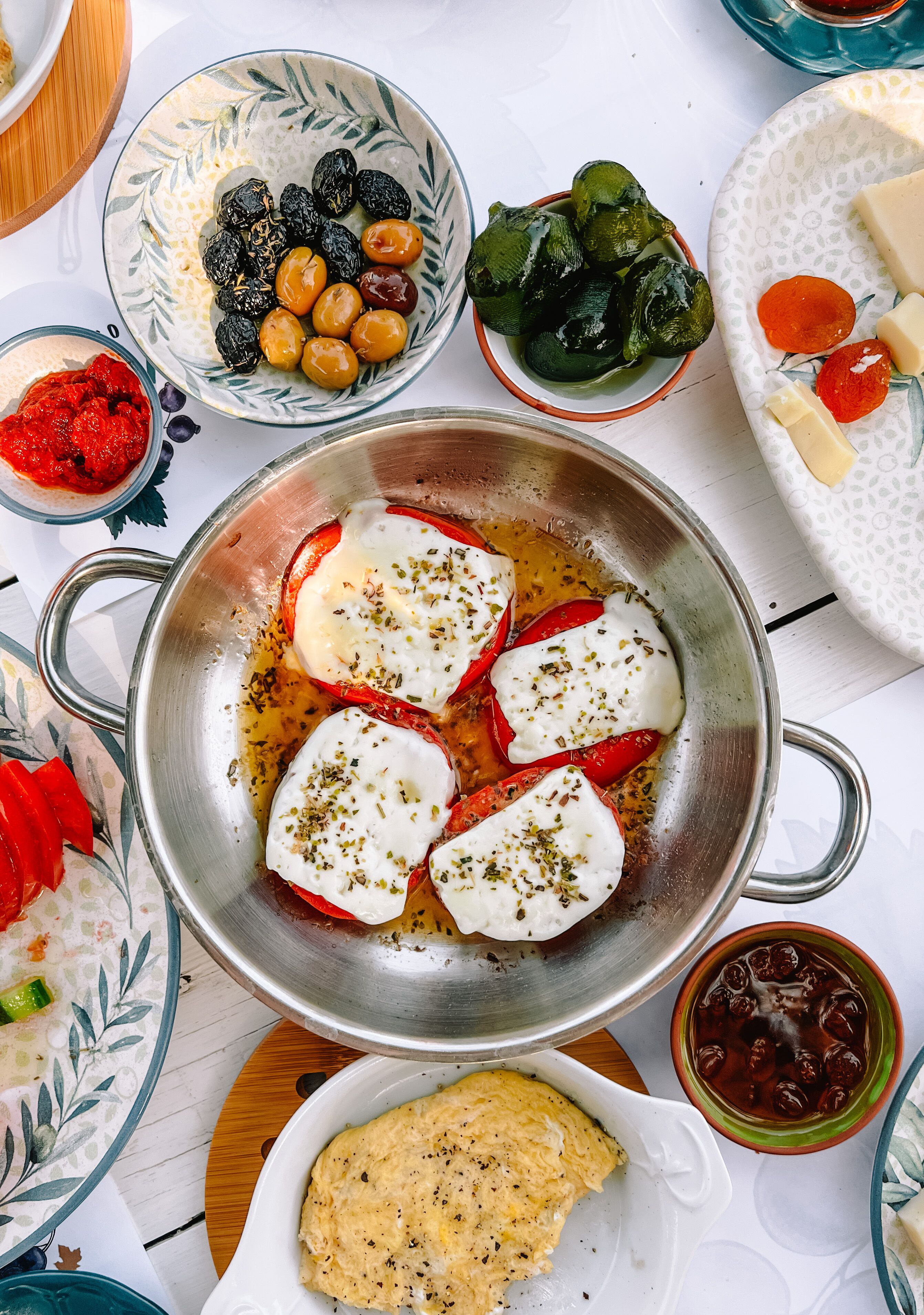 Diversos pratos, incluindo uma panela com queijo e tomate, tigelas de azeitonas e picles, e um prato com queijo e tomate.