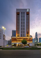 Um hotel moderno com entrada destacada, vários andares e paisagem urbana ao fundo.