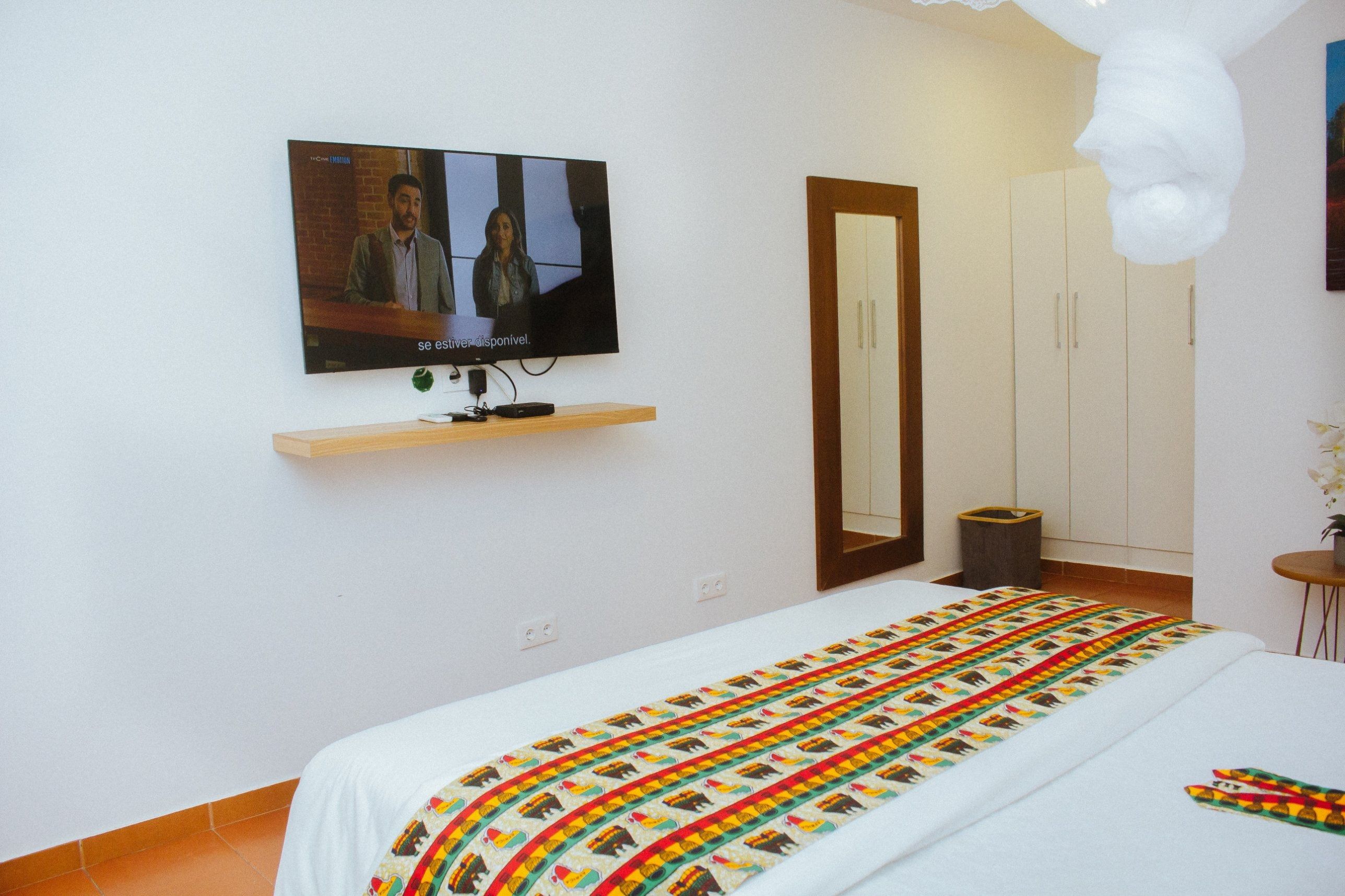 Quarto com cama, televisão na parede, espelho, guarda-roupa e uma pequena mesa com uma planta.