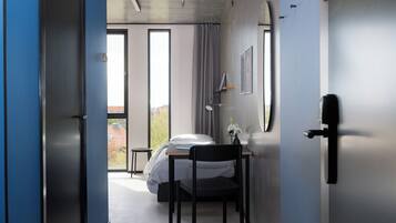 Quarto de hotel moderno com cama, escrivaninha e cadeira. Há uma janela com cortinas, uma porta e um candeeiro fixado na parede.