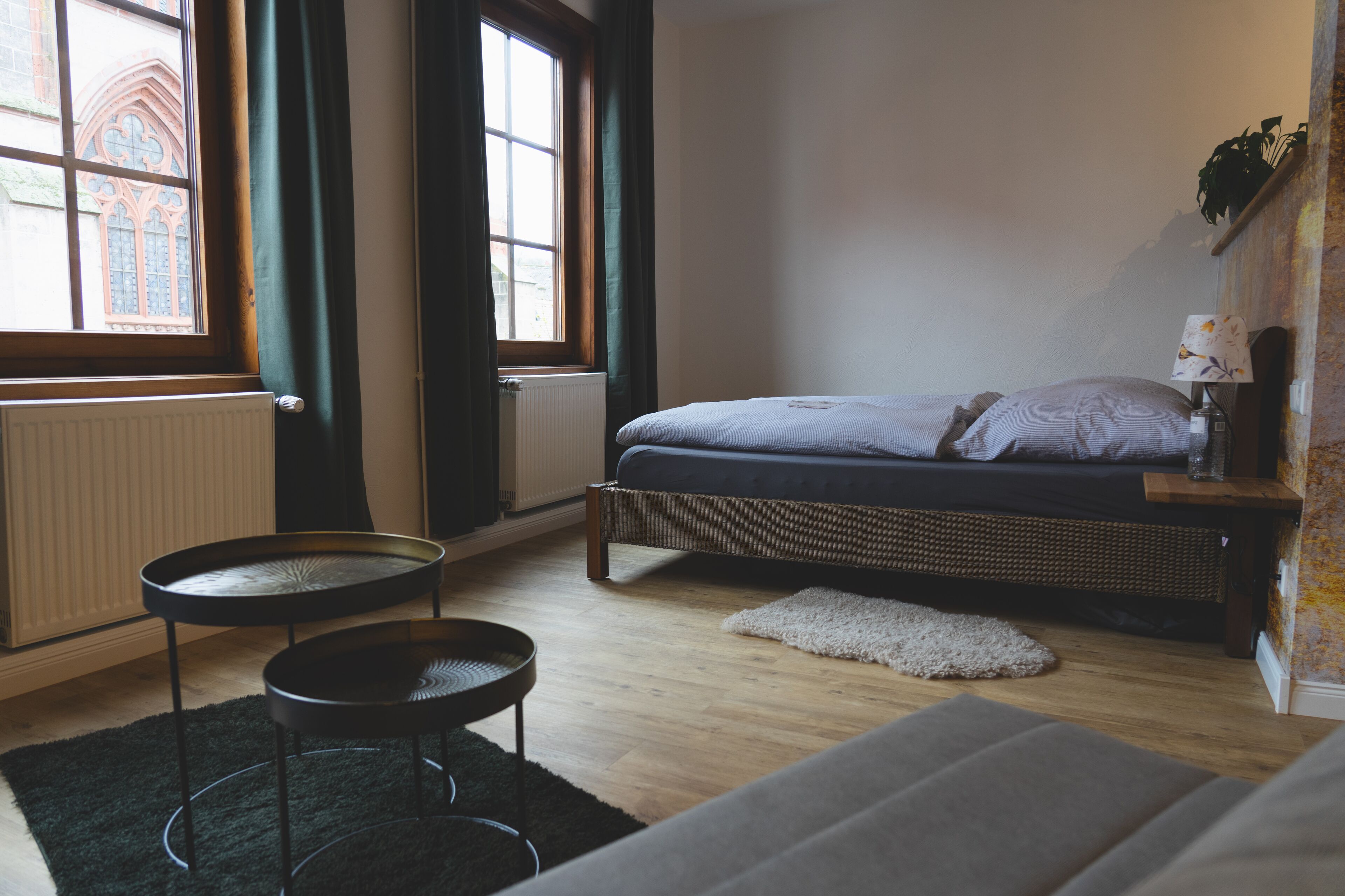 Quarto com cama, mesa redonda, radiador e janela com cortinas.