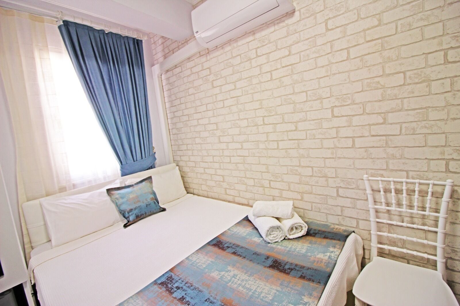 Quarto com cama, cadeira, janela com cortinas e parede de tijolos.