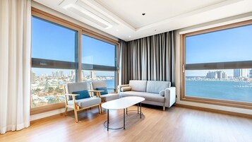 Quarto de hotel moderno com grandes janelas, um sofá, duas poltronas e uma pequena mesa.