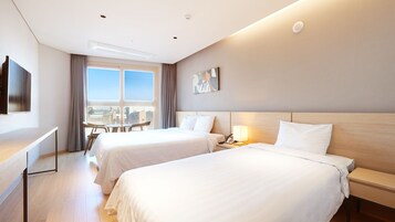 Quarto de hotel com duas camas, uma escrivaninha, uma cadeira, televisão e janela com vista para a cidade.