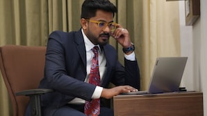 Um homem de terno escuro e gravata vermelha sentado em uma mesa com um laptop, em um escritório com cortina ao fundo.