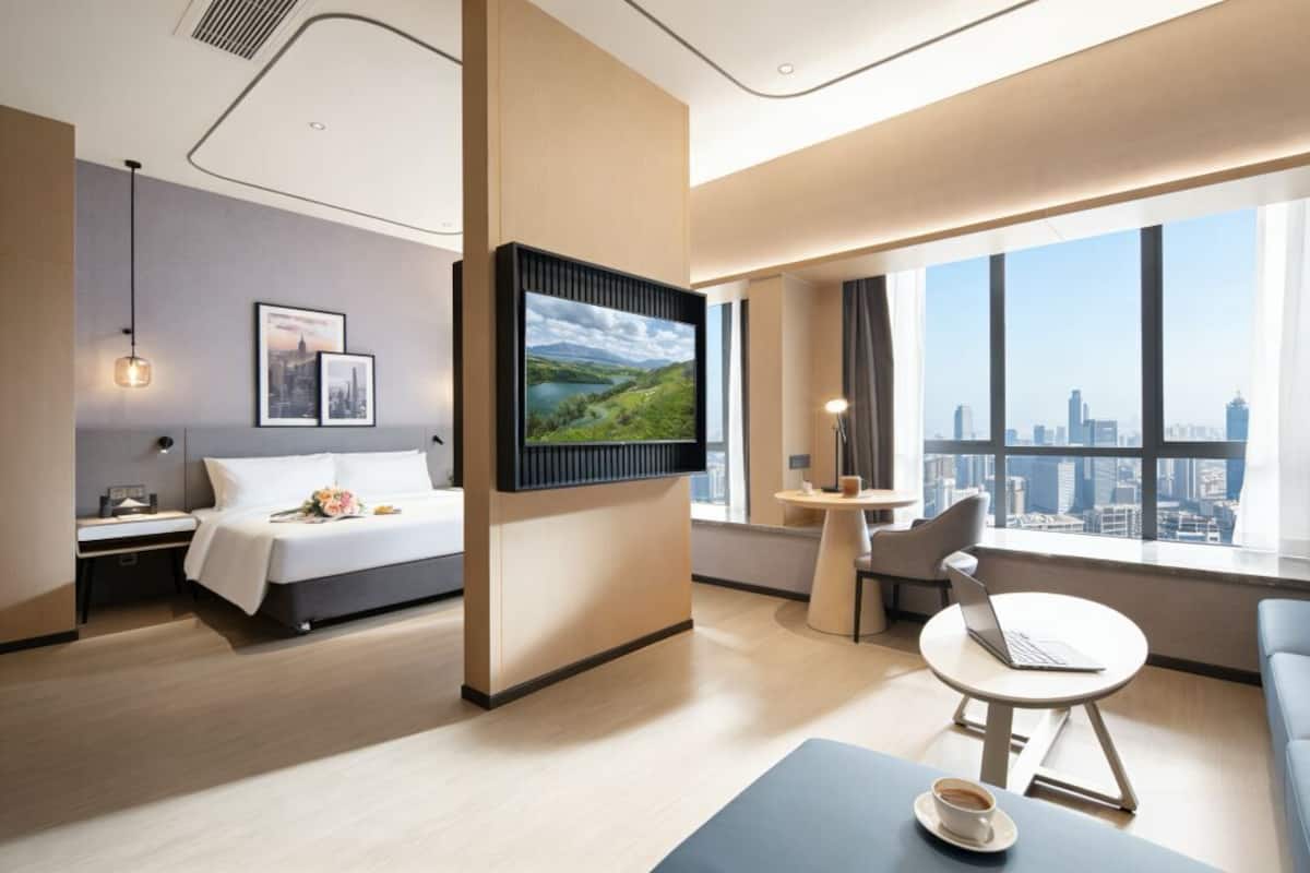 Quarto de hotel moderno com cama, escrivaninha, cadeira, laptop, xícara de café e uma grande janela com vista para a cidade.