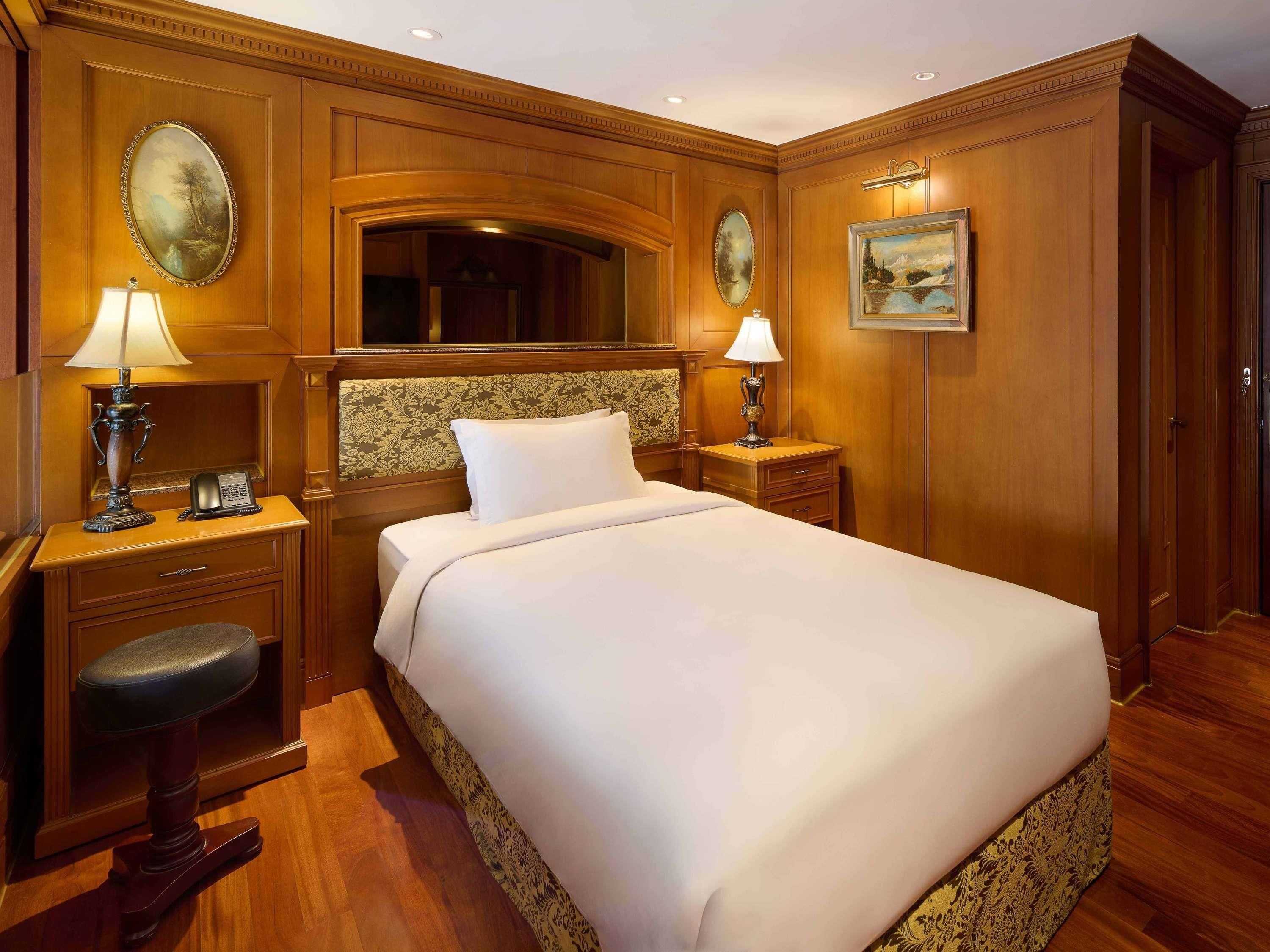 Quarto de hotel com uma cama grande, duas mesas de cabeceira com abajures, uma escrivaninha com telefone e uma cadeira.