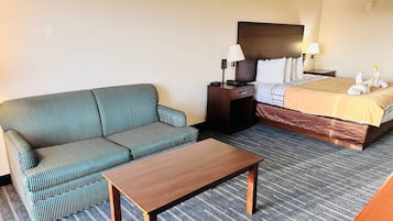 Quarto de hotel com um sofá listrado, uma mesa de madeira, uma cama com colcha amarela, uma mesa de cabeceira com abajur e uma televisão presa à parede.