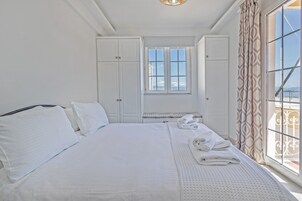 Quarto de hotel com cama grande, roupa de cama branca, janela com vista e varanda com cortinas estampadas.