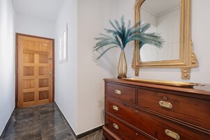 Um corredor com porta de madeira, chão de mármore, cômoda de madeira com gavetas, um vaso com penas azuis e um espelho com moldura dourada.