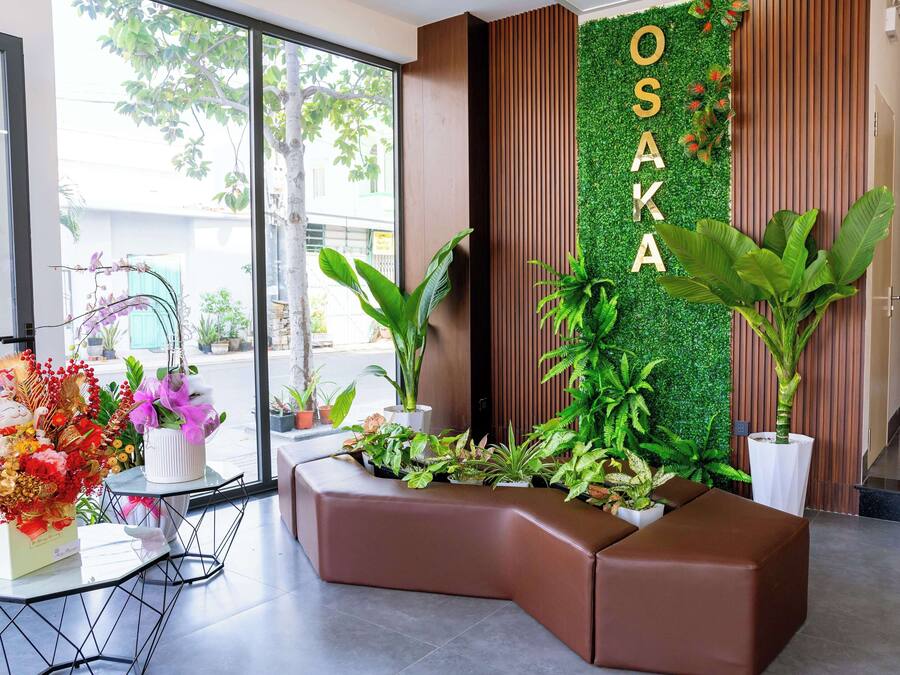 Uma área de recepção com um espaço de estar curvo em tons de marrom, uma parede verde com o nome 'OSAKA' e diversos vasos de plantas.