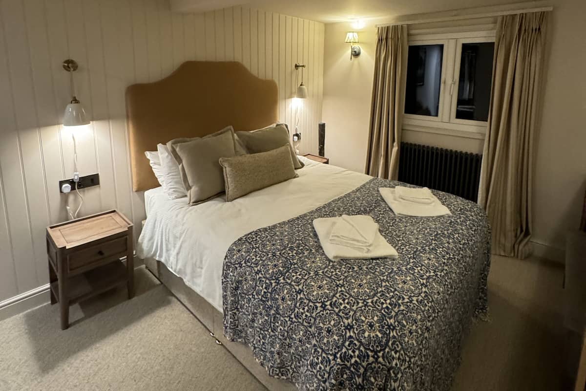 Quarto com cama, mesa de cabeceira, luminária e janela com cortinas.
