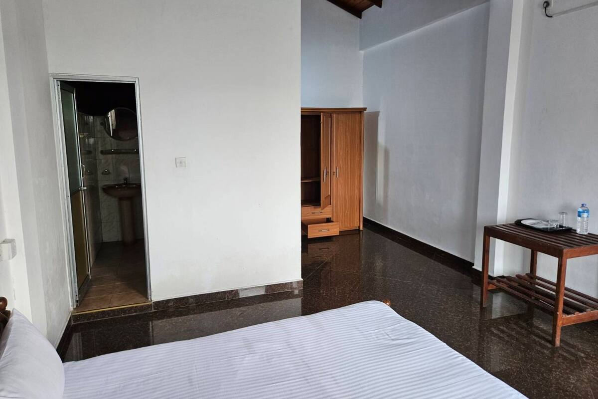 Quarto de hotel com uma cama, guarda-roupa de madeira, uma mesinha com uma garrafa de água e uma casa de banho com pia e espelho.