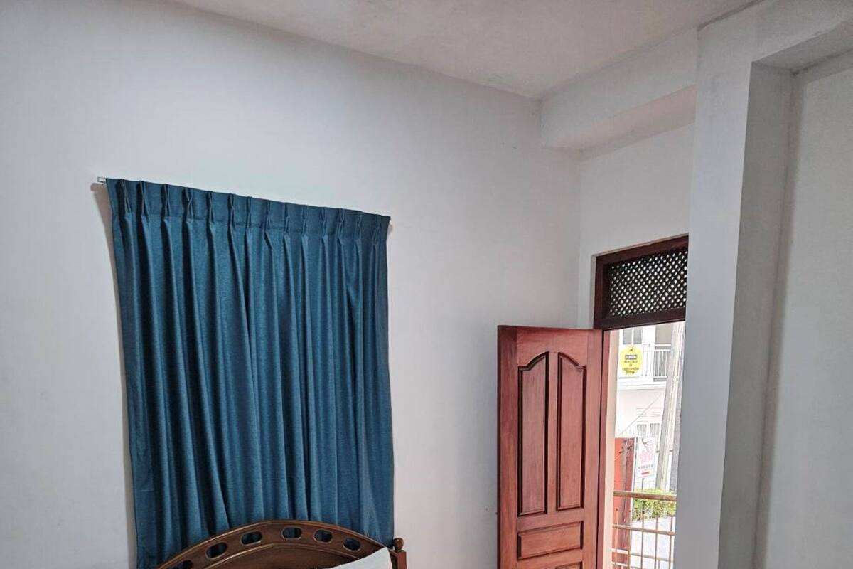 Uma cama de solteiro com cabeceira de madeira em quarto com soalho em azulejos e cortina azul.