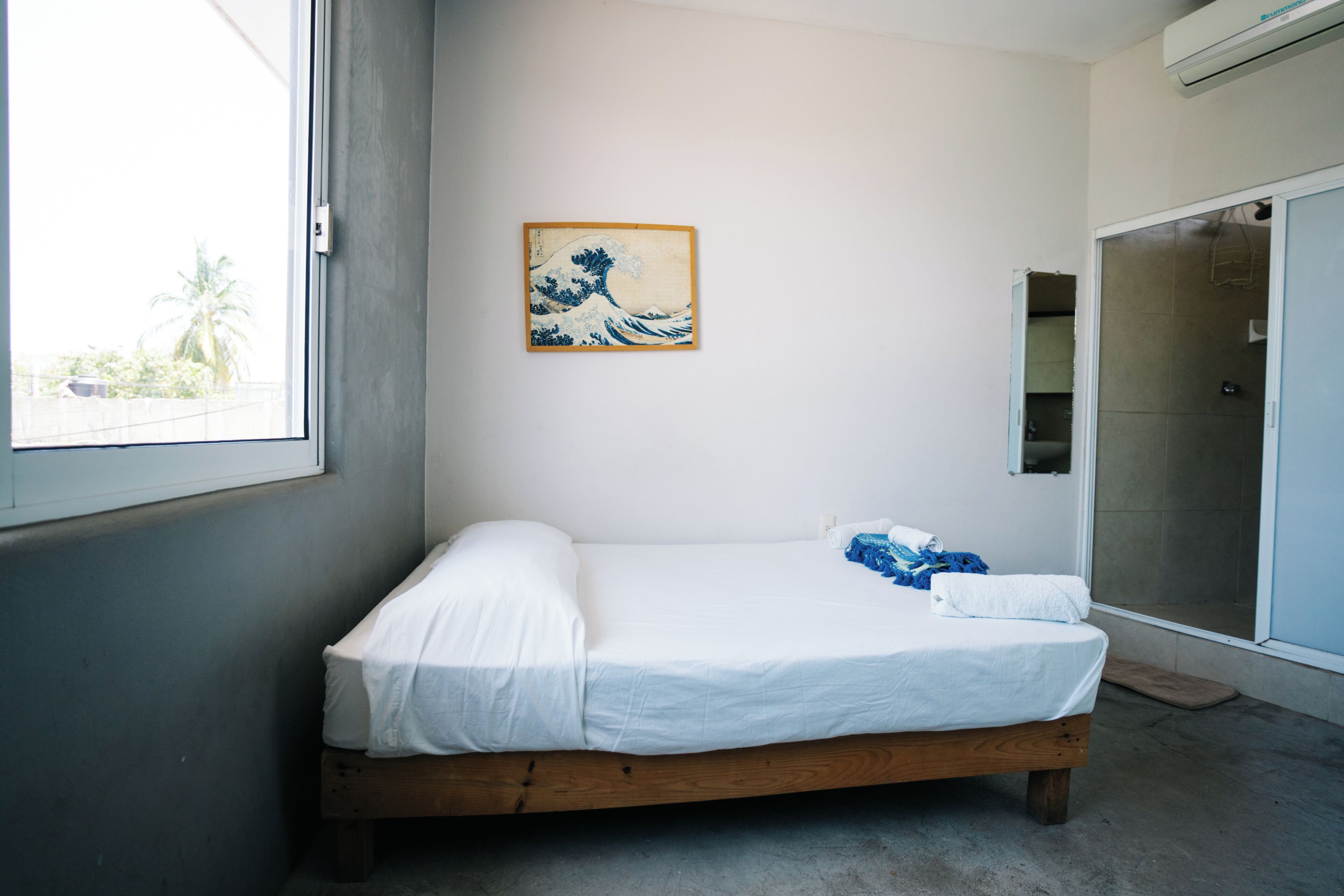Uma cama de casal com roupa de cama branca, cabeceira de madeira, janela com vista, uma obra de arte na parede e porta deslizante para área de chuveiro.