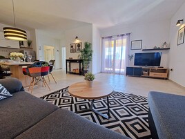 Uma sala de estar moderna com um sofá, uma mesa de centro e uma televisão. Há uma área de cozinha com uma geladeira e uma mesa de jantar.