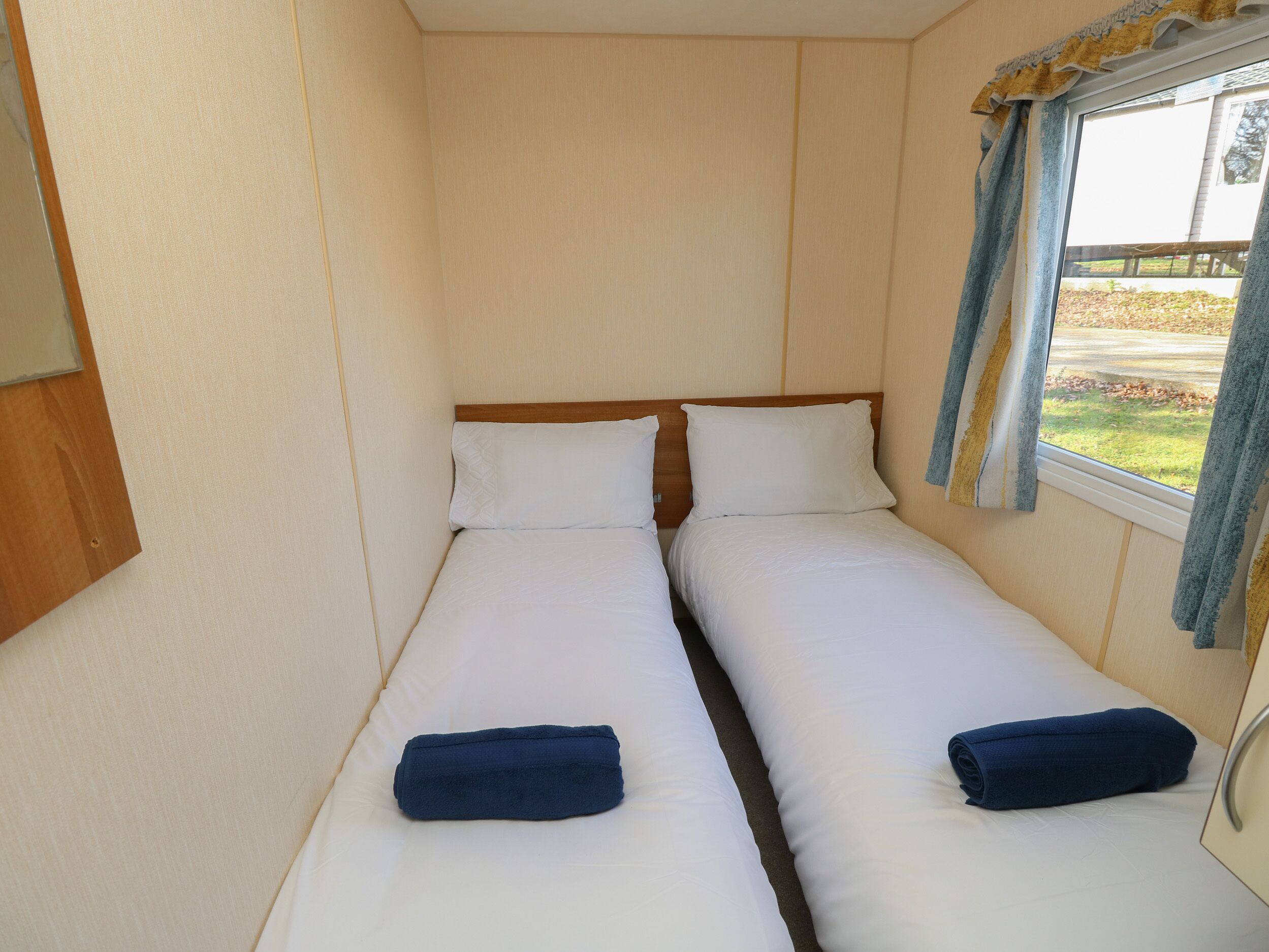 Uma cabana pequena, com um quarto e duas camas, uma janela com cortinas e uma cabeceira de madeira.