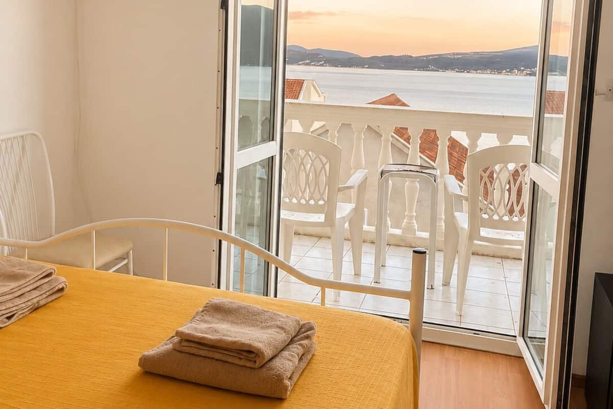 Quarto com cama, mesa com toalhas, televisão e varanda com cadeiras brancas e vista para o mar.