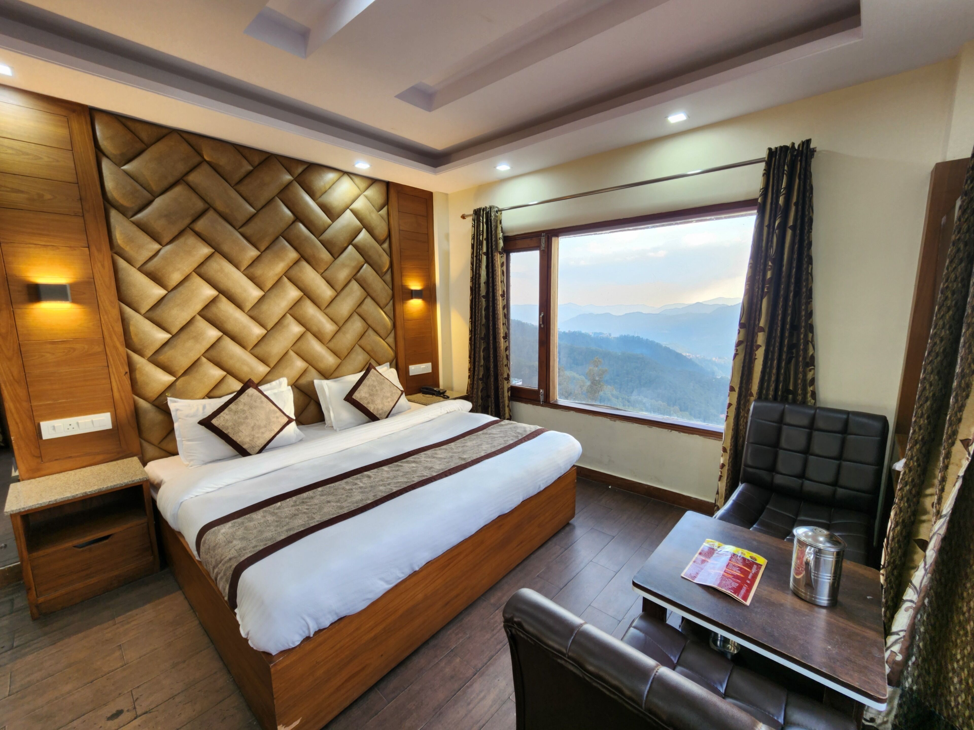 Quarto de hotel com uma cama grande, cabeceira de madeira, vista para as montanhas, uma mesa pequena com uma revista e um termo, e uma poltrona de couro.