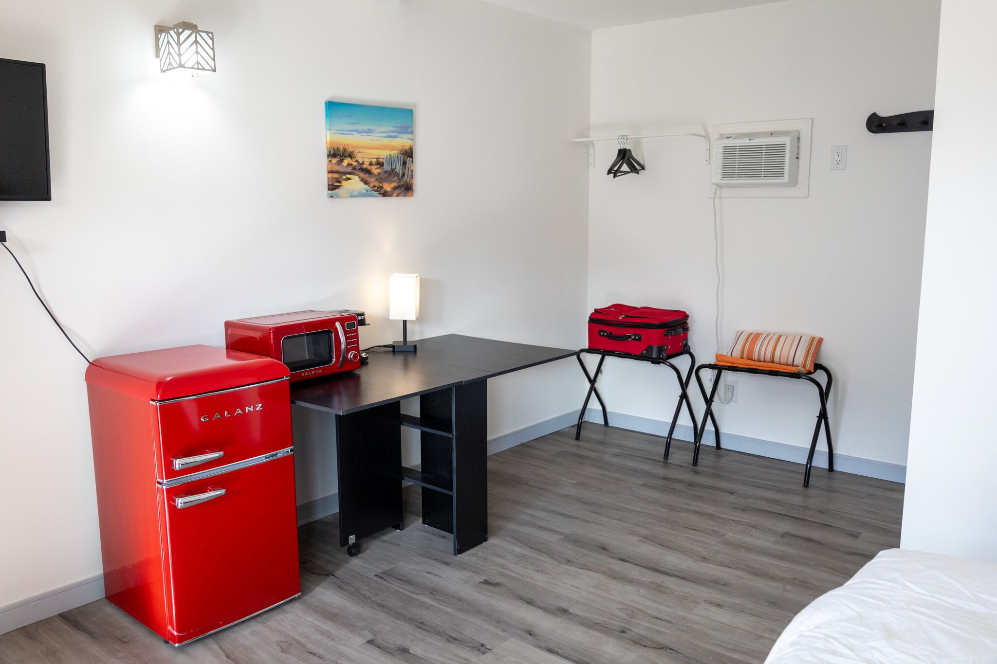 Quarto compacto com cama, escrivaninha com luminária, geladeira vermelha, mala vermelha e uma cadeira.