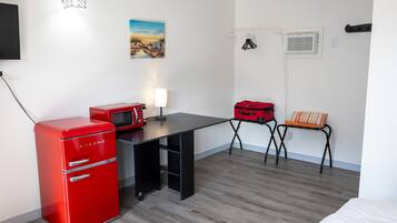 Quarto compacto com cama, escrivaninha com luminária, geladeira vermelha, mala vermelha e uma cadeira.