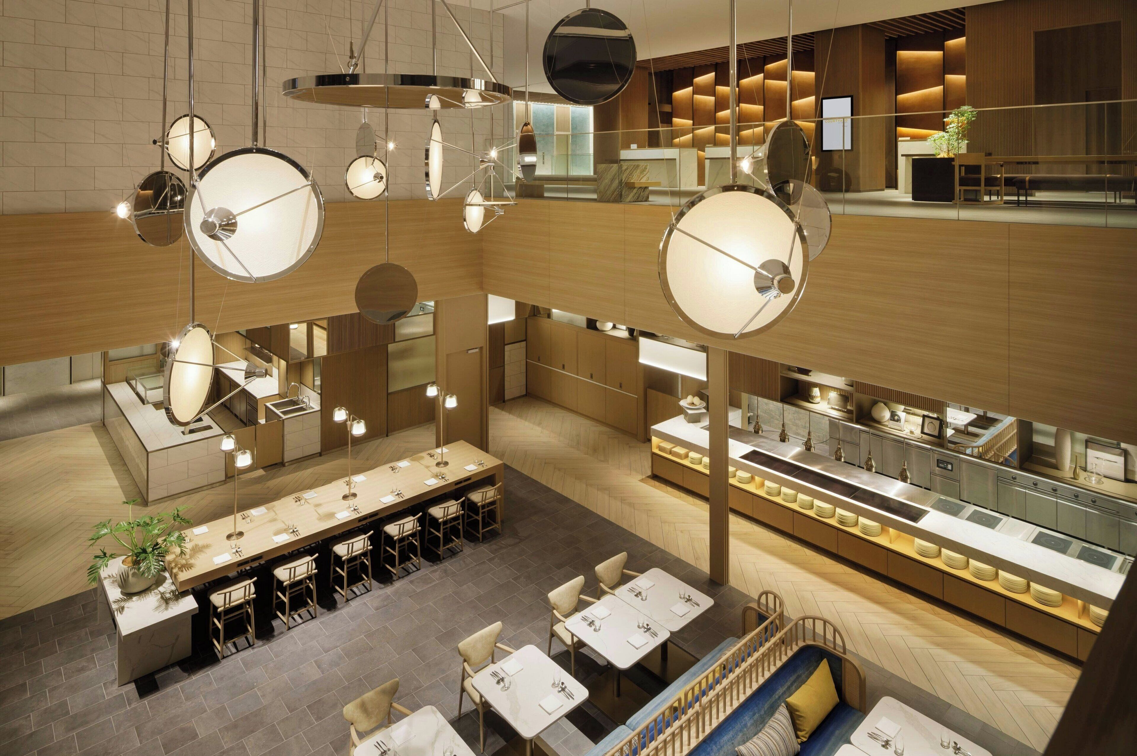 Um hall de hotel moderno com bar central, diversas áreas de refeição e um amplo átrio com altos tetos e luminárias esféricas penduradas.