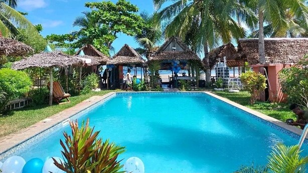 Piscina azul límpida rodeada por palmeiras e estruturas com telhados de palha, provavelmente parte de um resort.