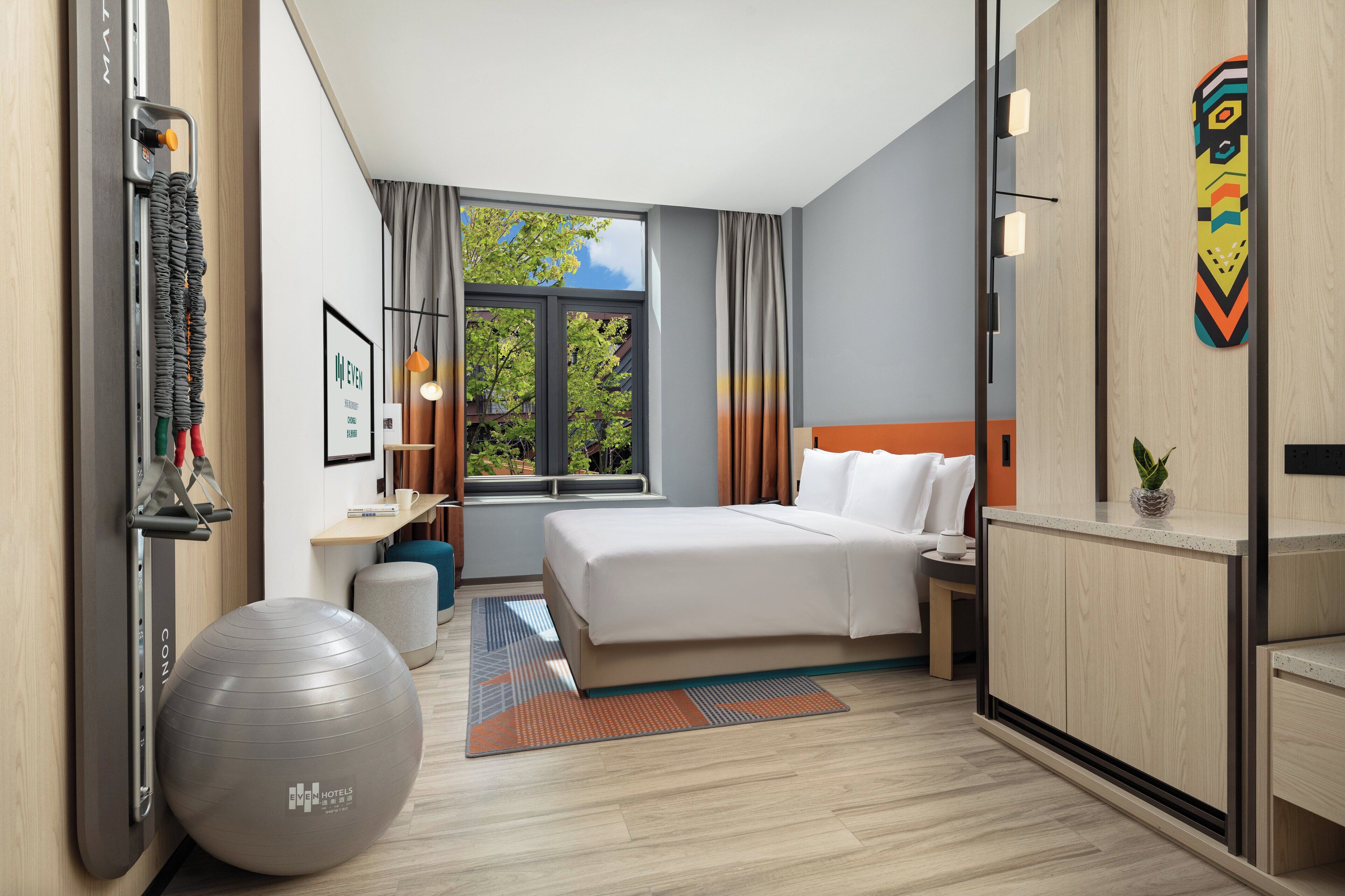 Quarto de hotel com cama, bola de exercícios, escrivaninha e janela com cortinas.
