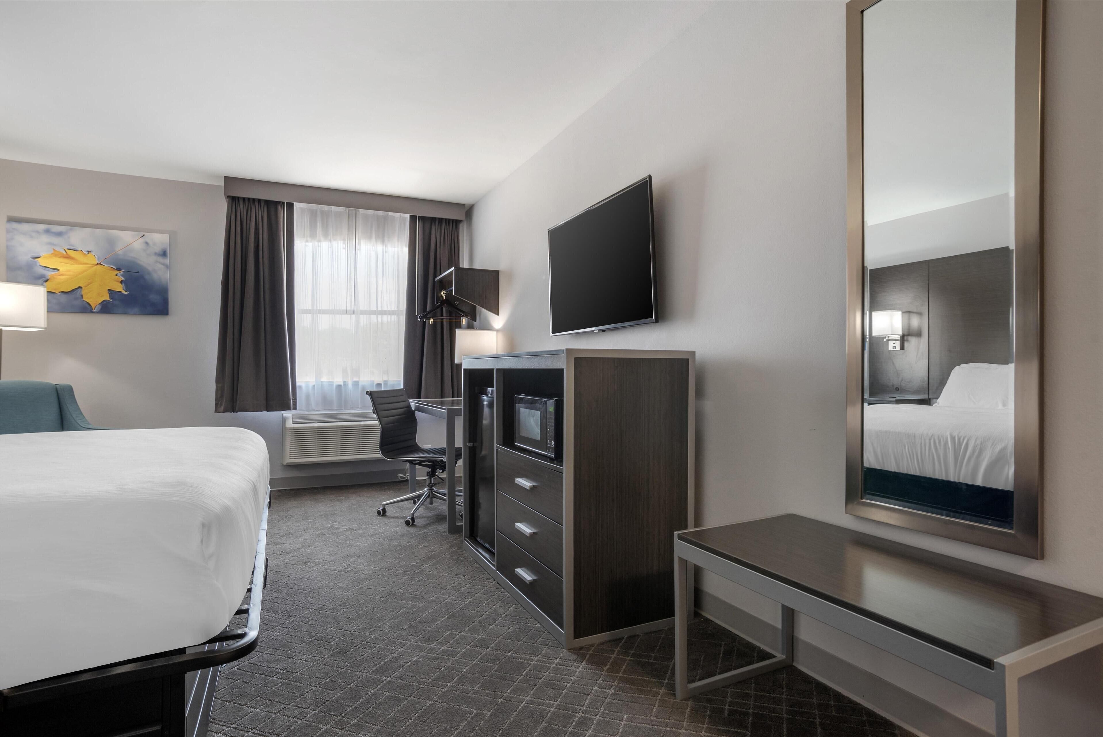 Quarto de hotel com cama, escrivaninha, televisão, espelho, cadeira, abajur e janela com cortinas.